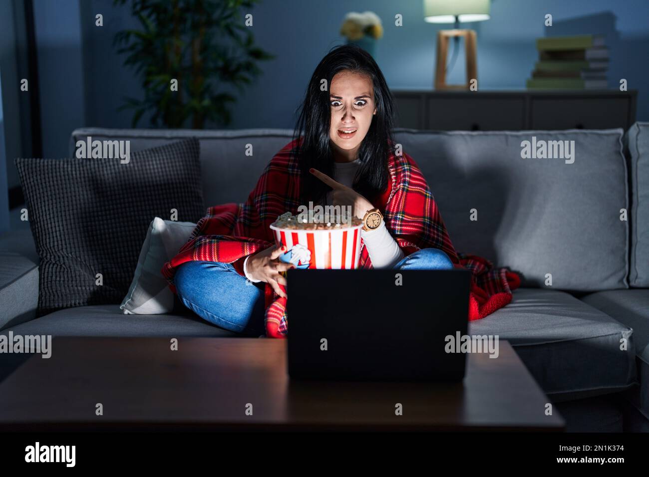 Hispanic woman eating popcorn watching a movie on the sofa pointing ...