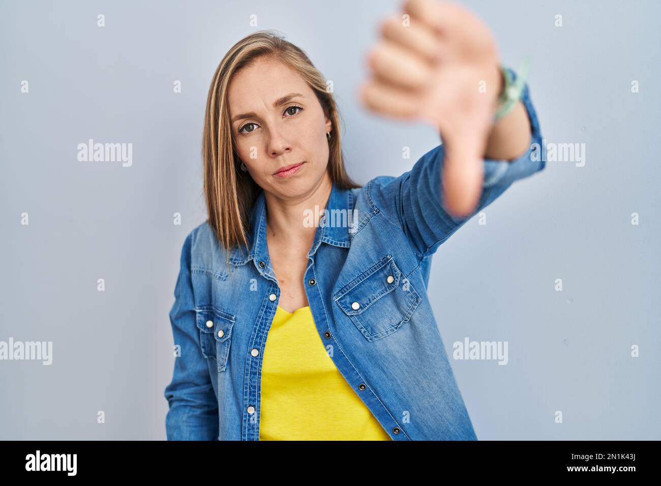 Young blonde woman standing over blue background looking unhappy and ...