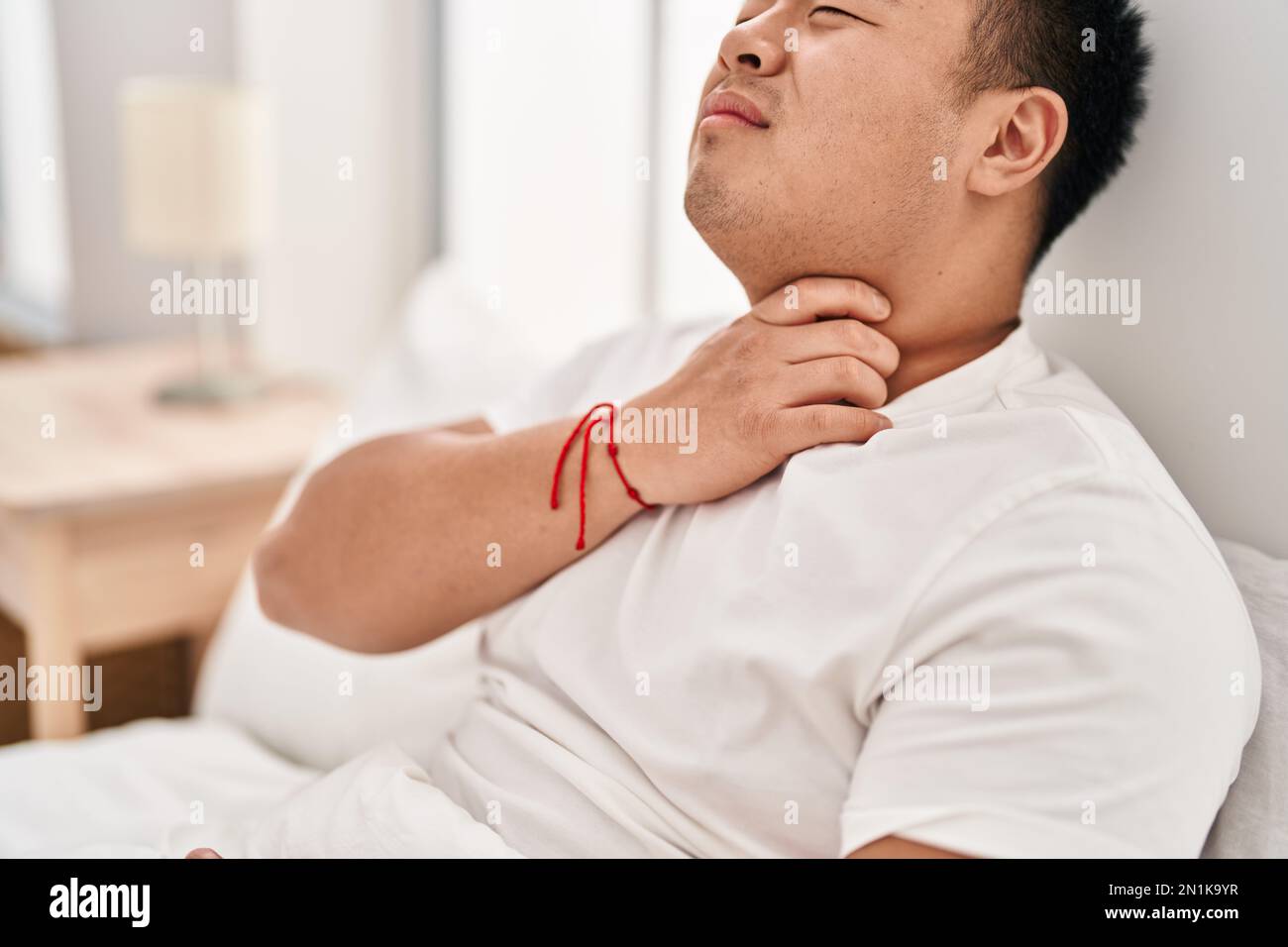 Young chinese man suffering for neck pain sitting on bed at bedroom ...