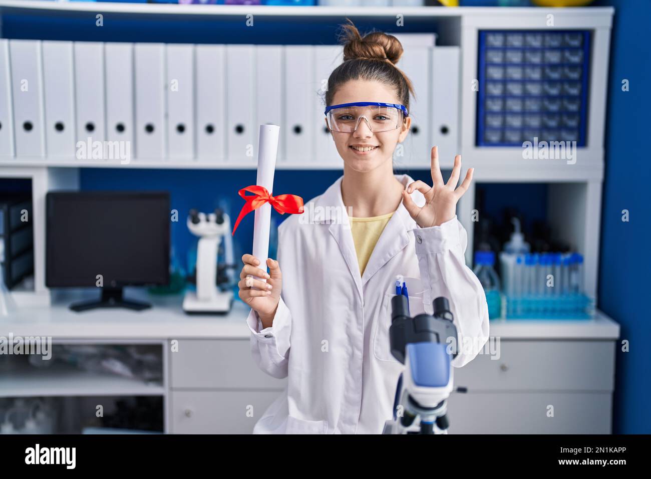 Teenager girl working at scientist laboratory holding degree doing ok ...