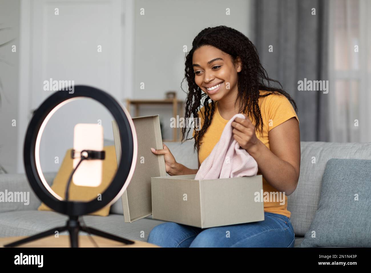 Happy black female influencer unpacking box with clothes and recording video on cellphone camera ...