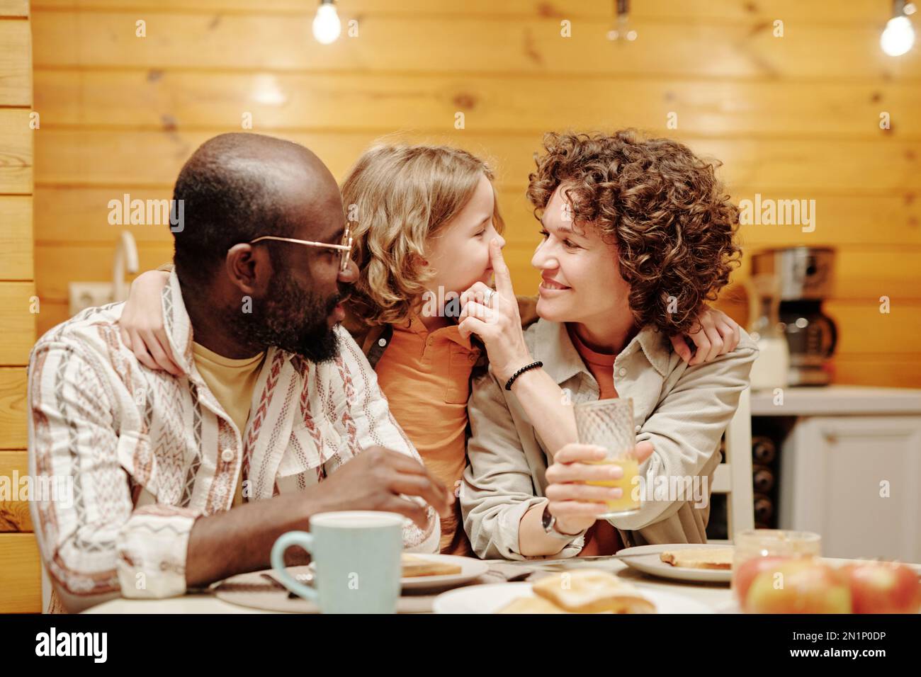 Cute little boy embracing his happy intercultural parents by breakfast ...