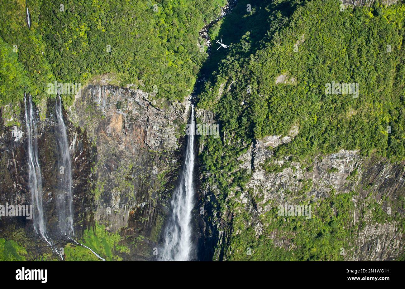 A helicopter passes over a waterfall on the forested lower slopes of ...