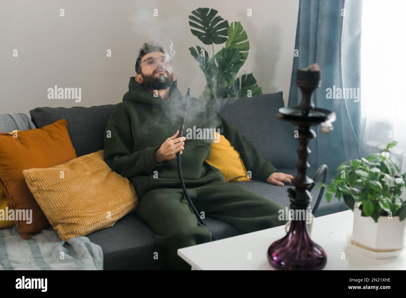 Bearded millennial or gen z man smoking hookah while relaxing on sofa ...