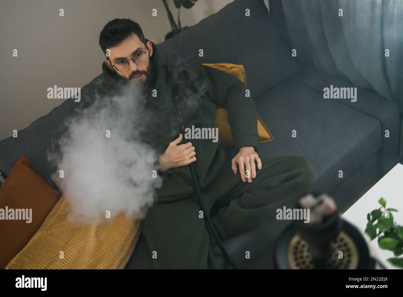Bearded millennial or gen z man smoking hookah while relaxing on sofa ...