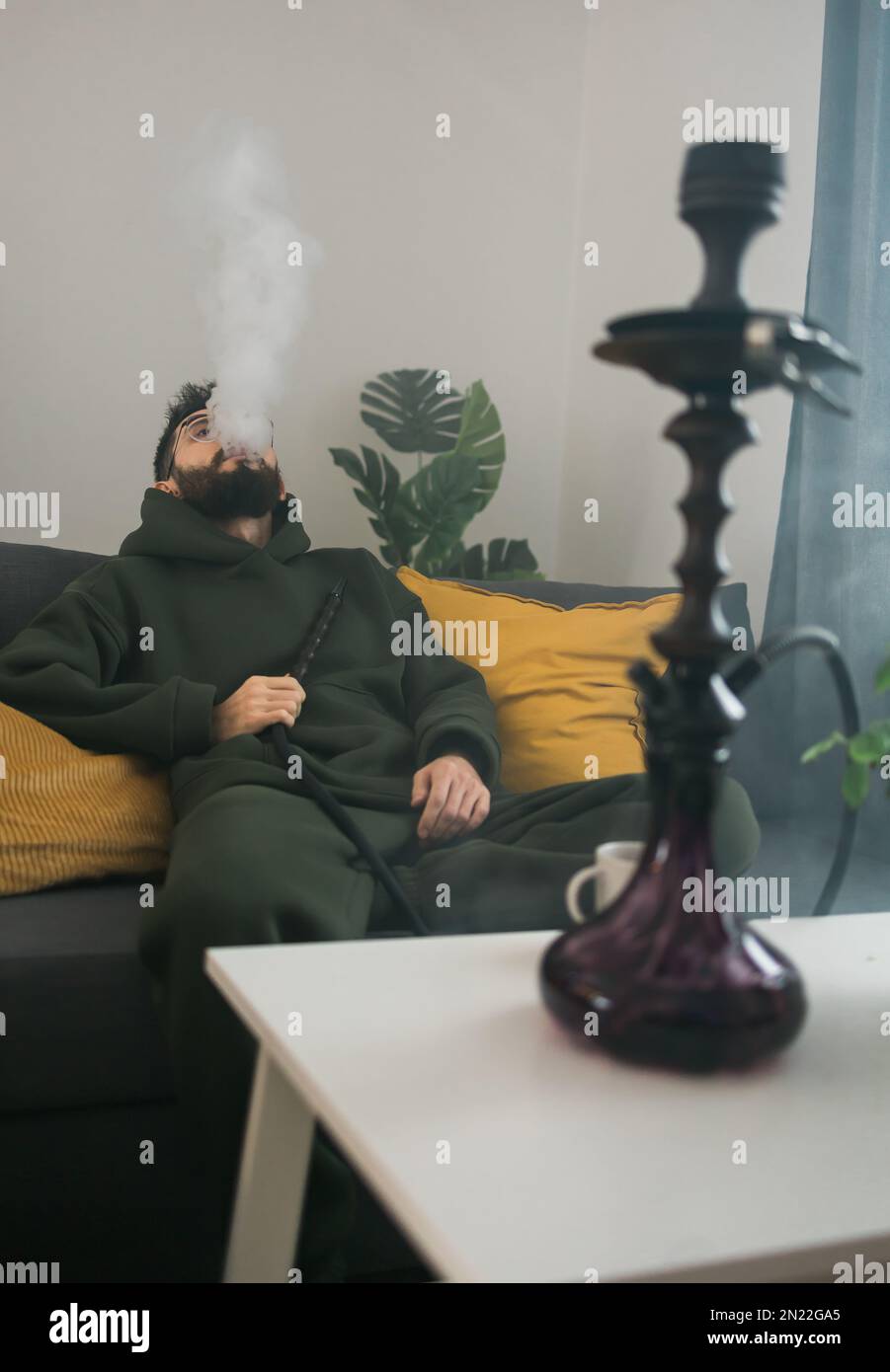 Bearded millennial or gen z man smoking hookah while relaxing on sofa ...