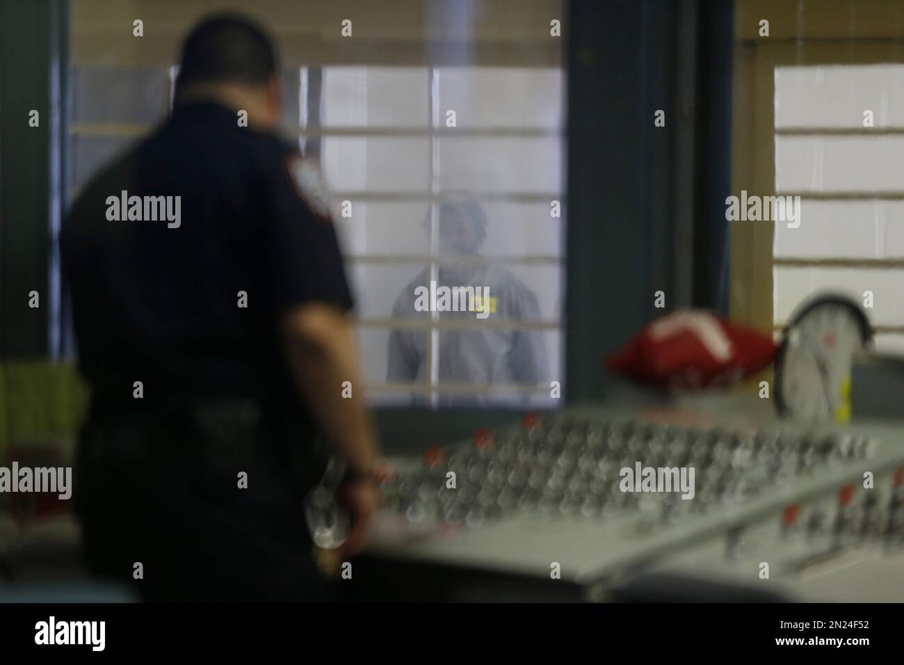 A prisoner behind bars and thick plastic looks over at a corrections ...