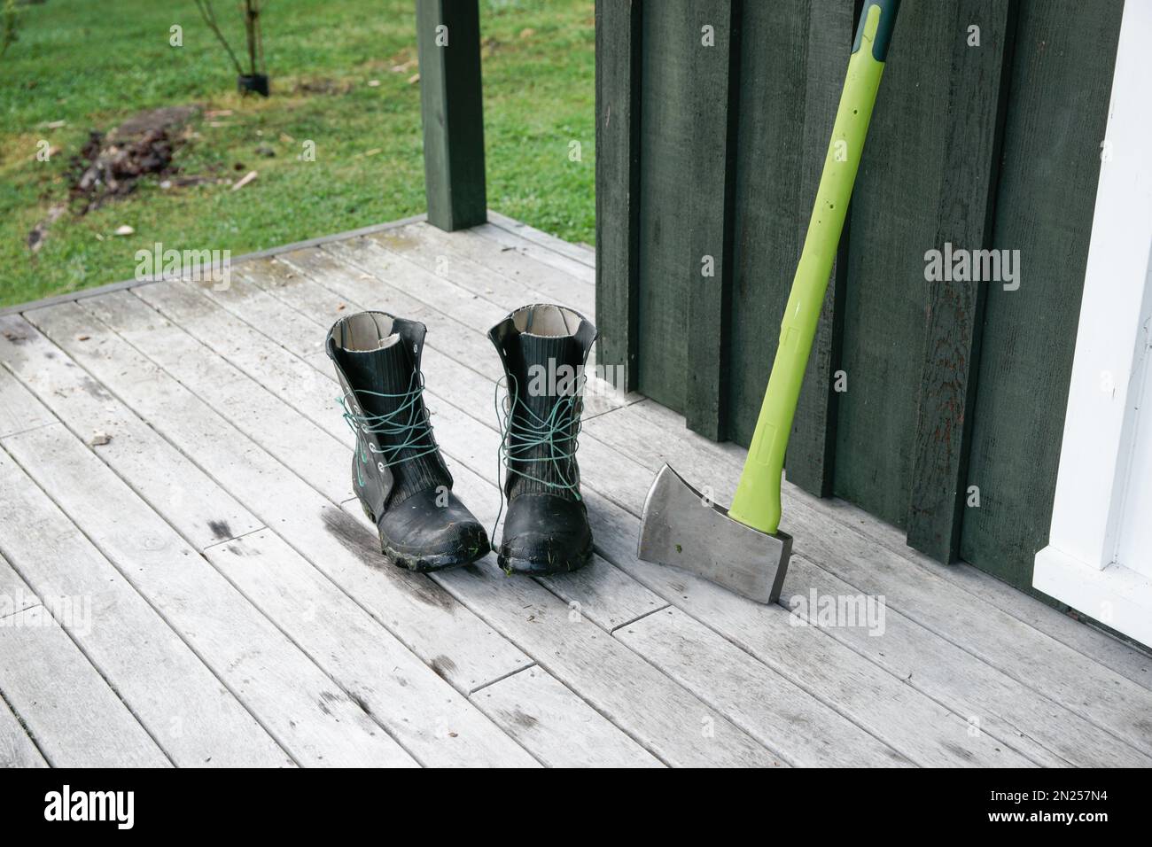 Old black boots and axe with bright green axe handle on deck of shed ...