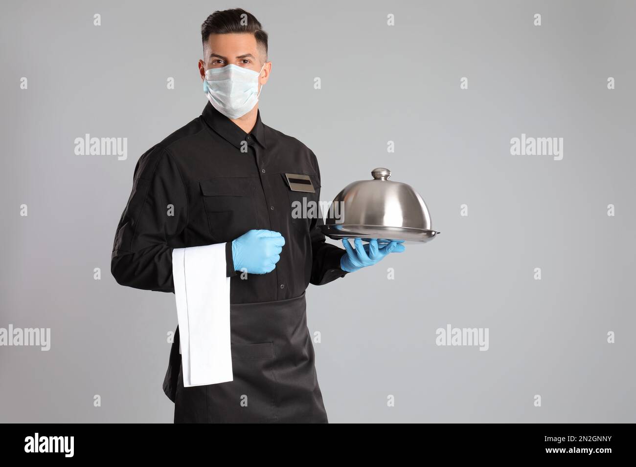 Waiter in medical face mask holding tray with lid on light grey ...