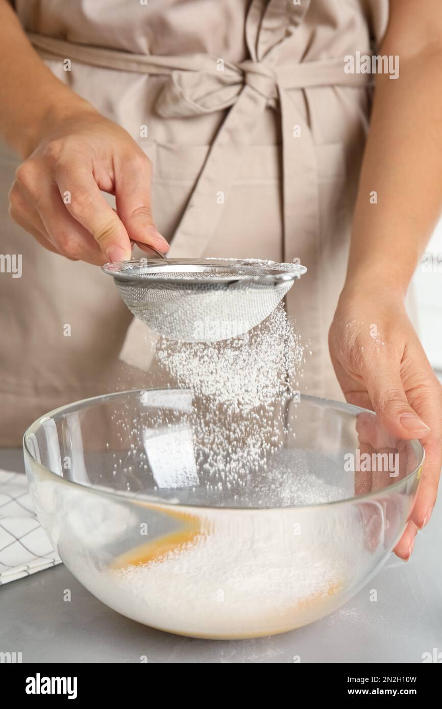Woman sieving flour into bowl at light grey table, closeup. Cooking of ...
