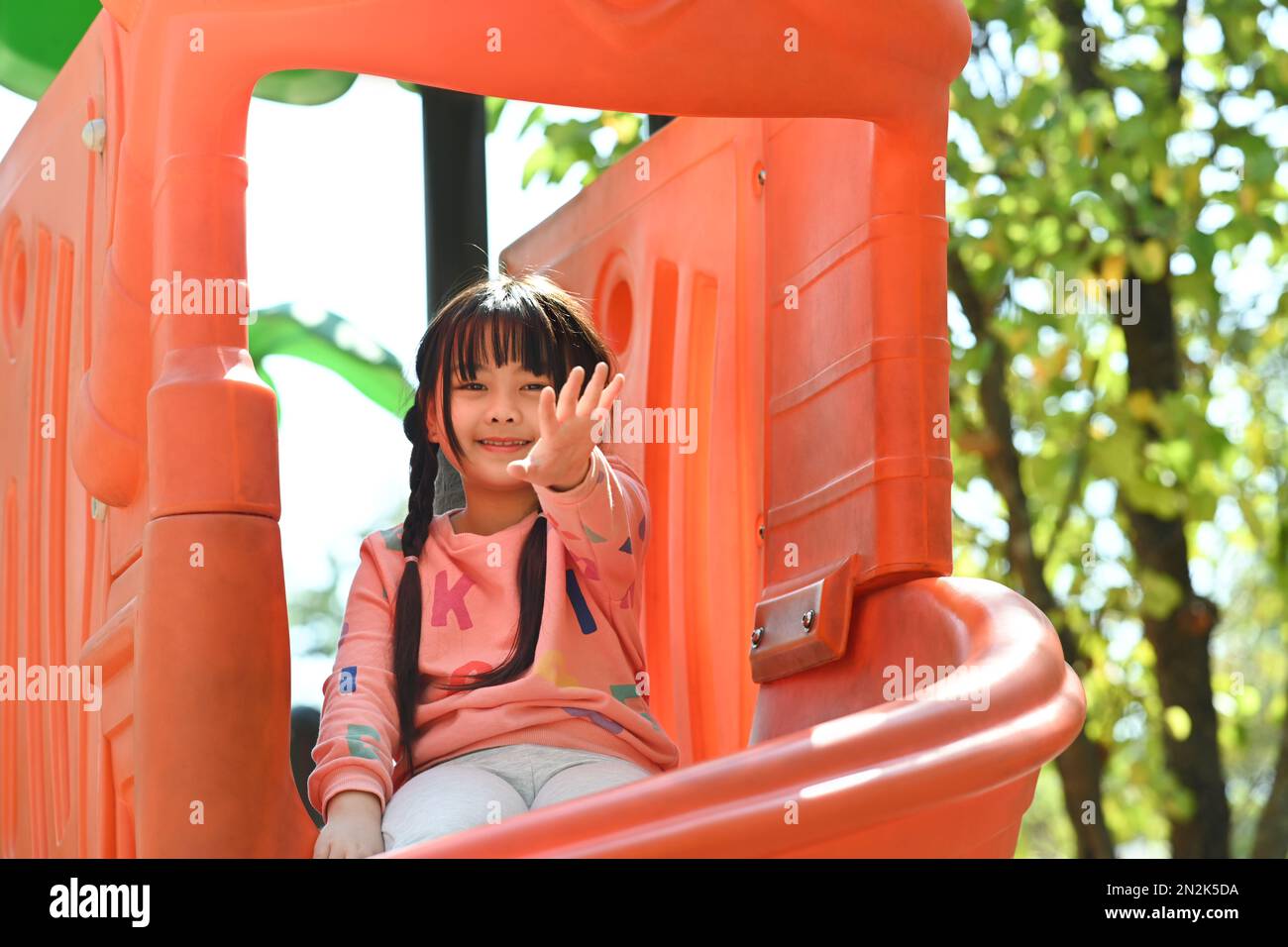 Happy Little Asian Girl Having Fun On Playground In Public Park Surrounded By Green Trees At