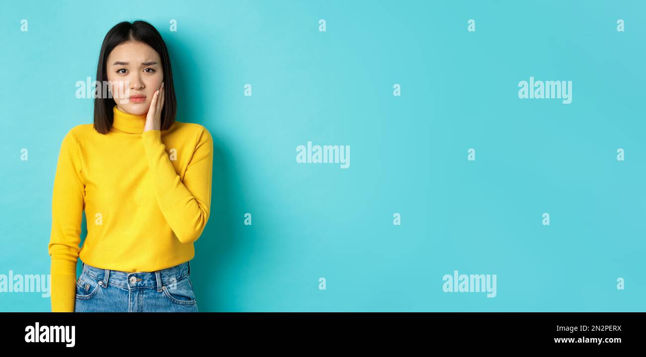 Portrait of asian young woman touching cheek and frowning, looking sad ...
