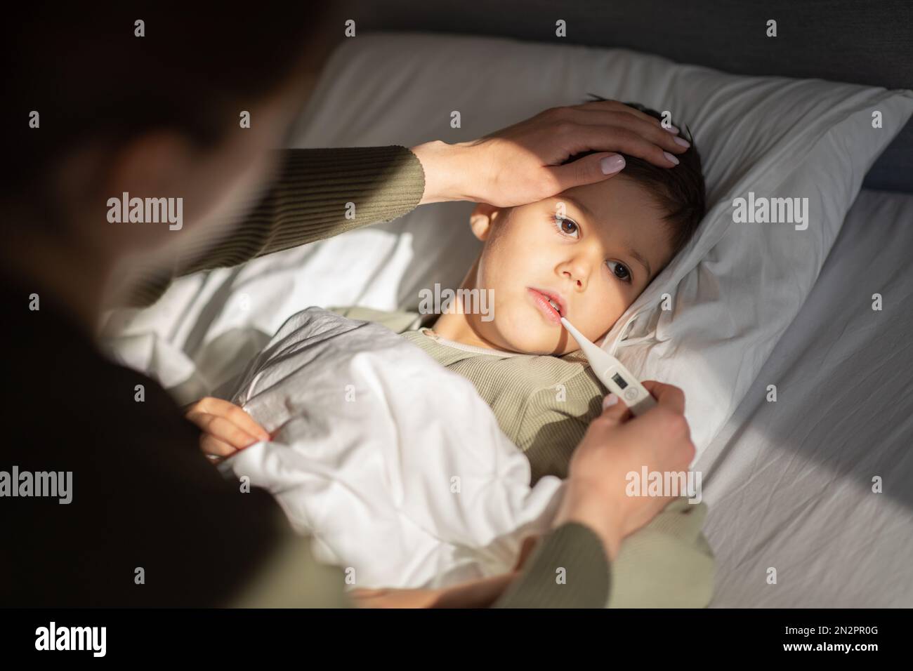 Unhappy caucasian little child suffers from fever, mom measures high temperature with ...