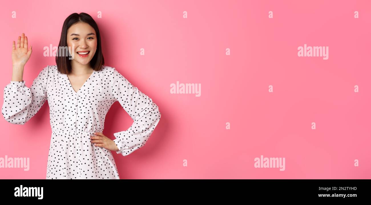 Beautiful asian woman in dress saying hello, waving hand to greet and ...