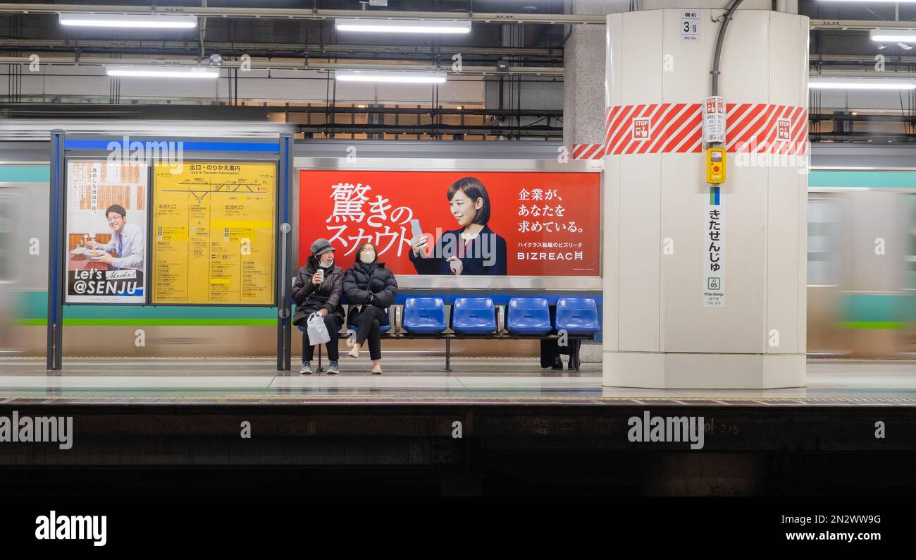 Two middle-aged ladies talking while sitting on a train platform bench waiting in the cold for ...