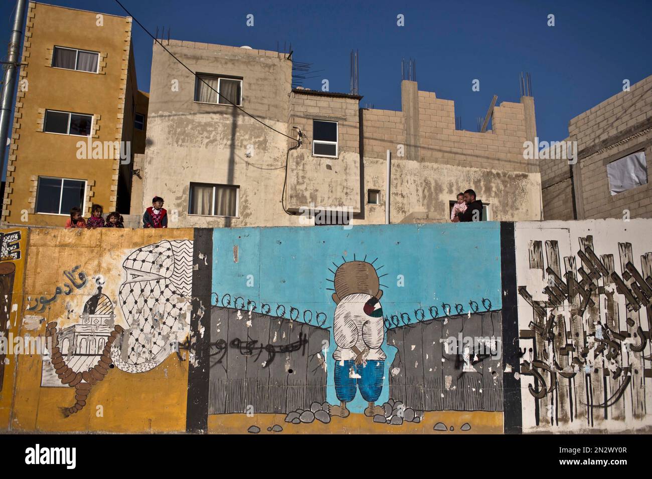 Palestinian refugees walk behind a wall painted with a mural depicting ...
