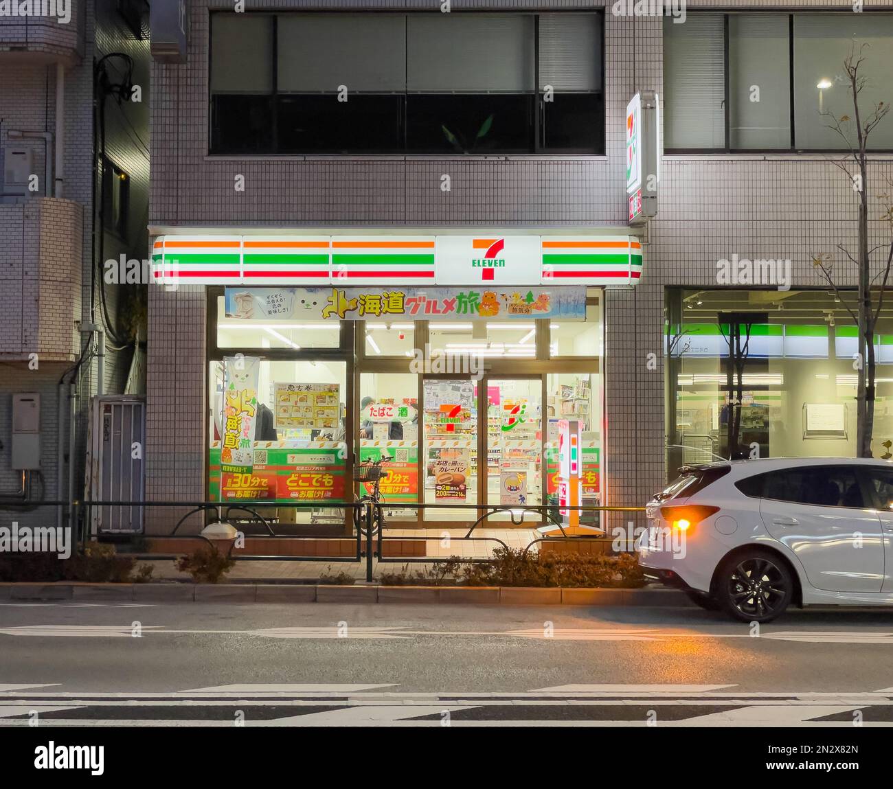 A bright storefront shot of a busy 7-Eleven convenience store at dusk with lots of buildings ...