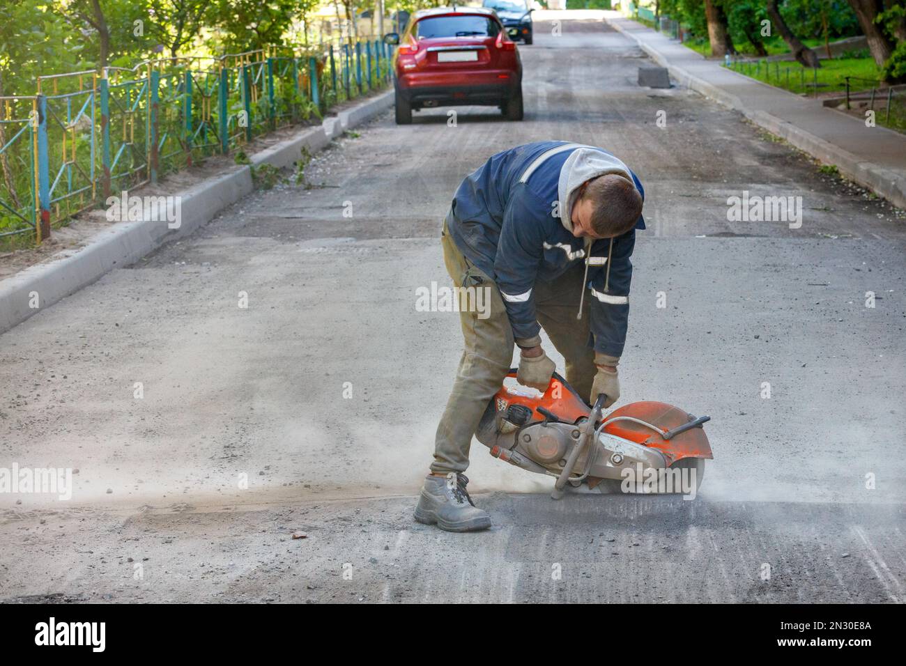 A worker cuts worn-out asphalt with a petrol cutter in a cloud of dust ...