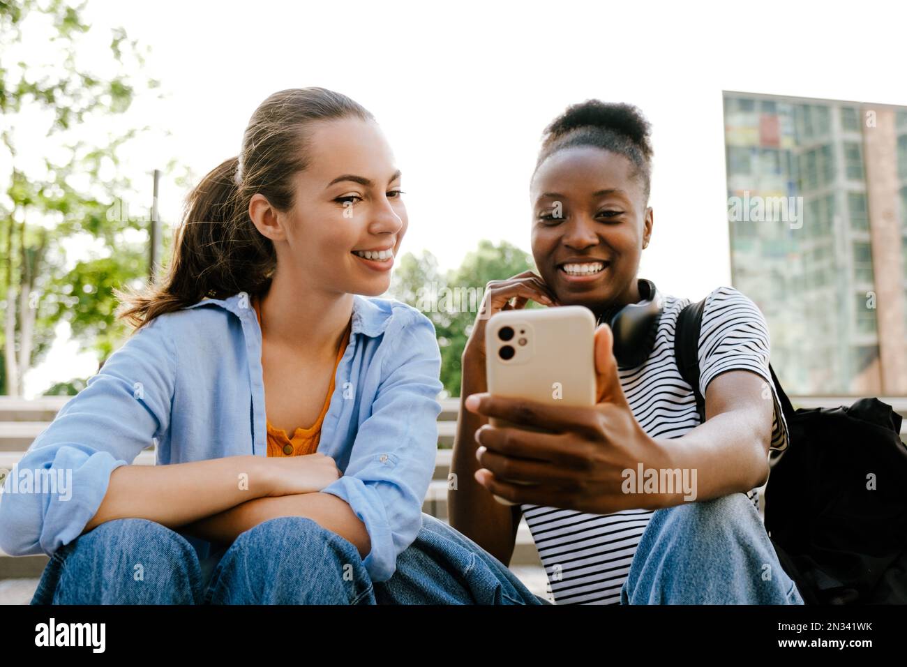 Young multinational girls talking and using cellphone together while sitting on stairs outdoors ...