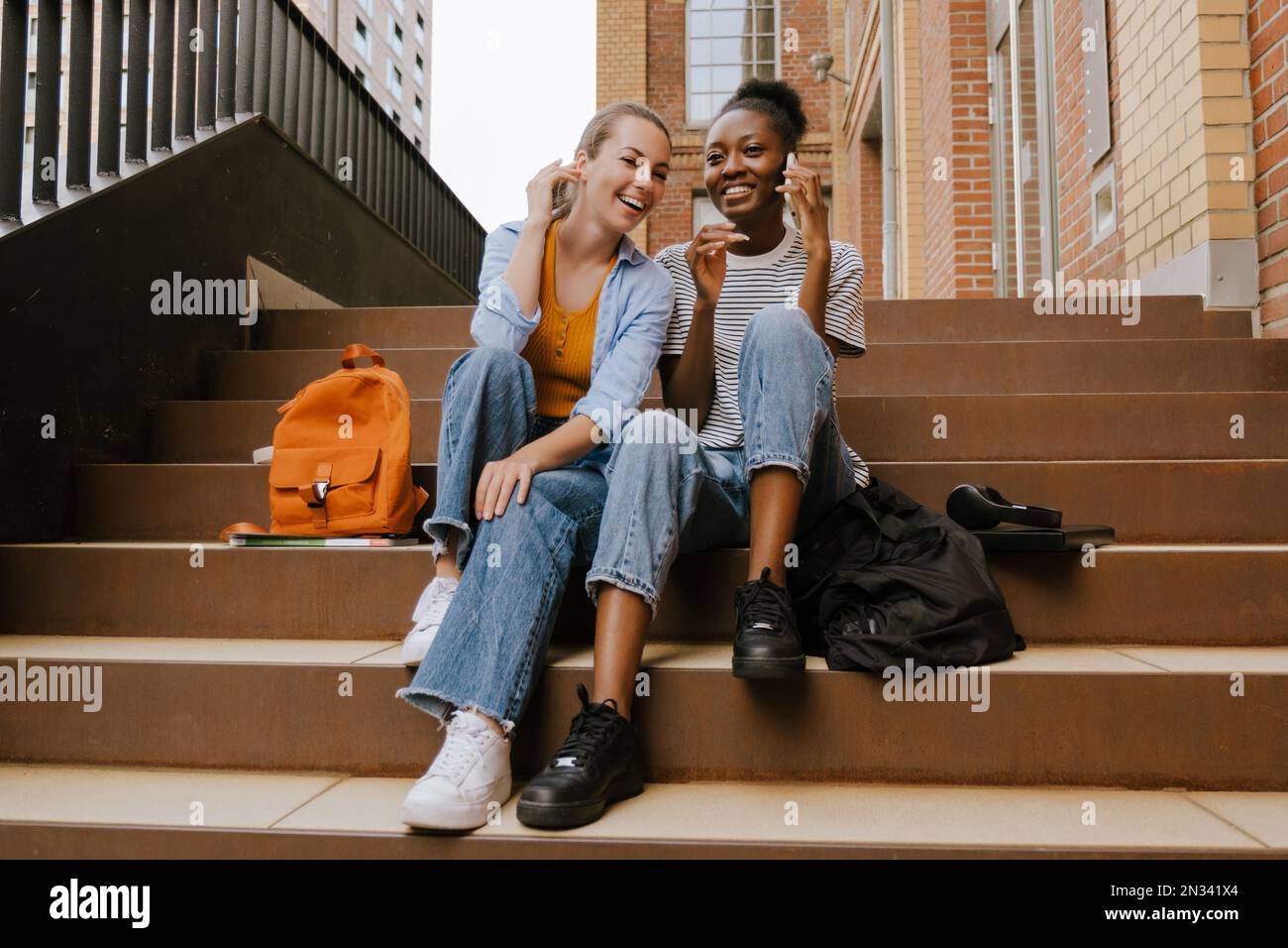 Young multinational girls smiling and using cellphone together while sitting on stairs outdoors ...