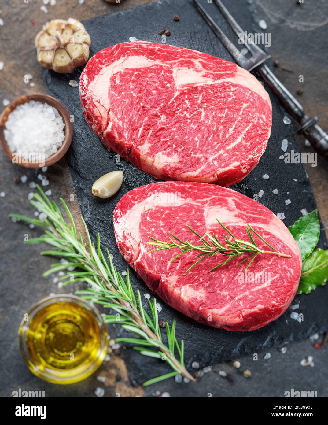 Raw ribeye steaks with rosemary and other seasoning on gray tray. Top ...