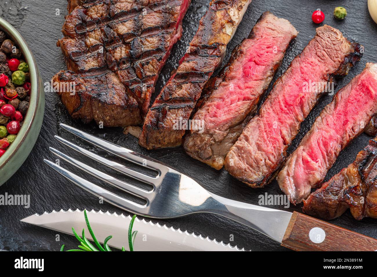 Grilled delicious ribeye steak slices and food utensils on black slate ...