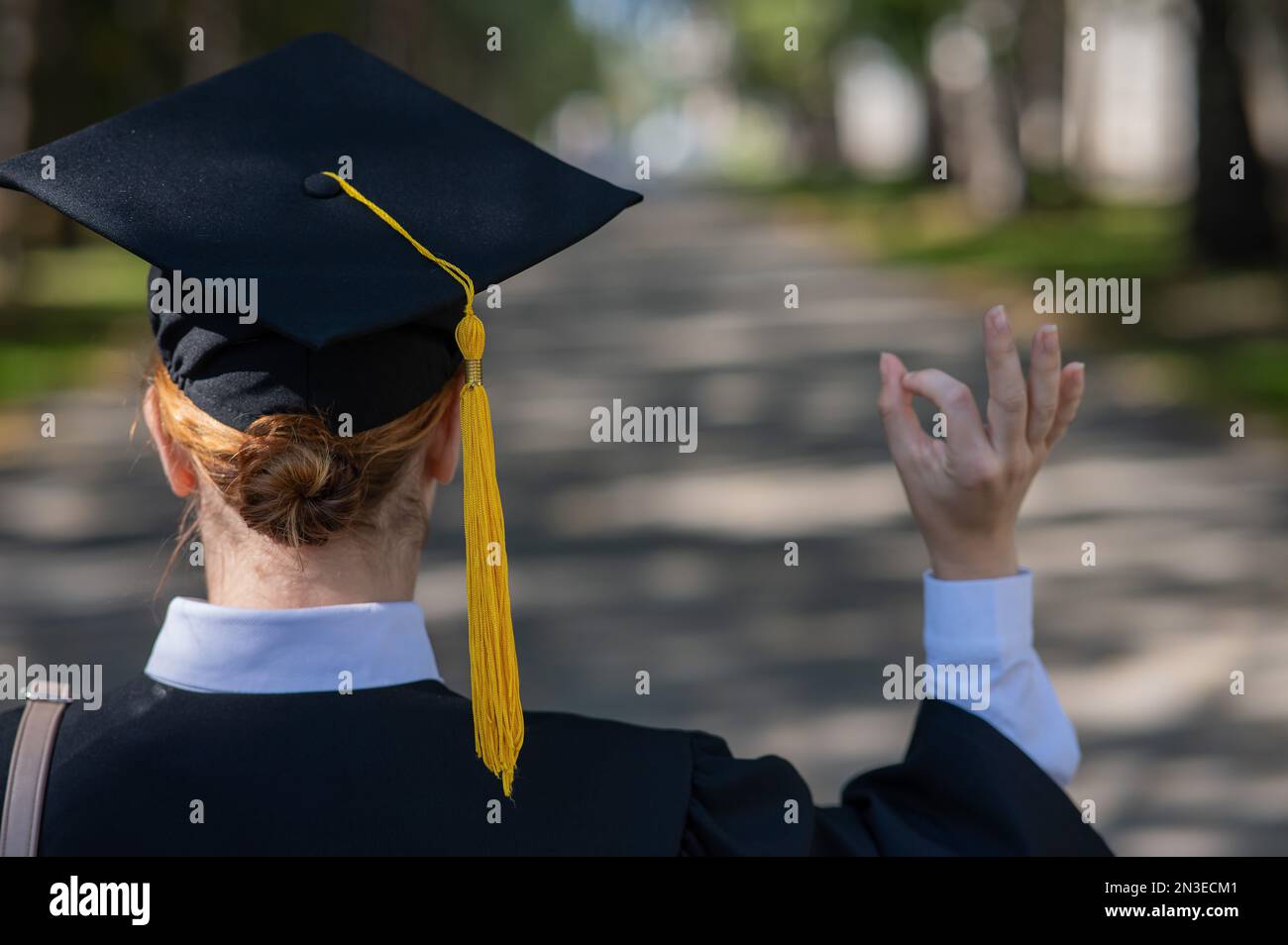 Rear view of caucasian woman in graduate gown showing ok sign outdoors ...