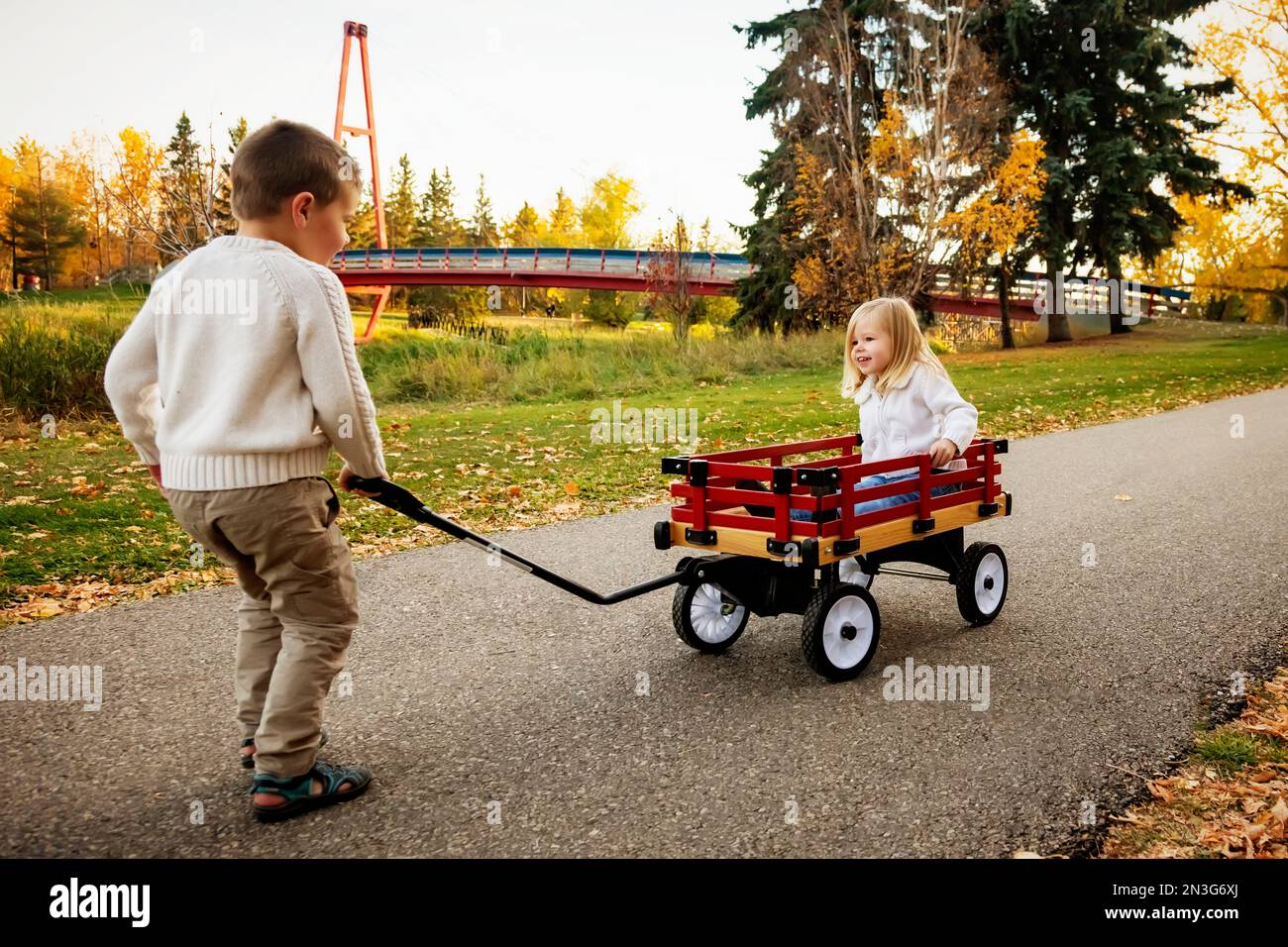 Young boy pulling his sister in a wagon at a city park along a river ...
