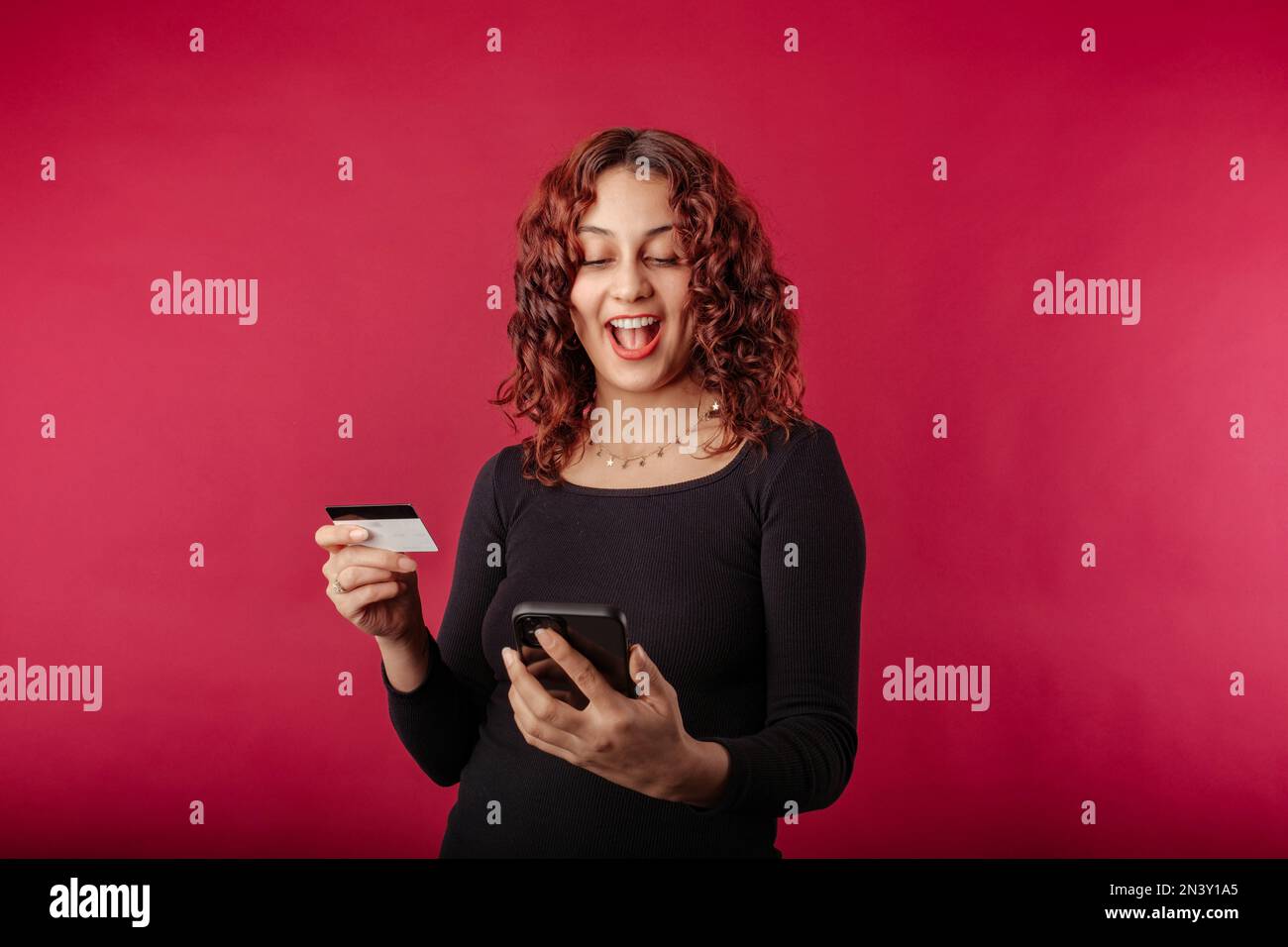 Happy redhead woman standing isolated over red background holding a credit card and makes a ...
