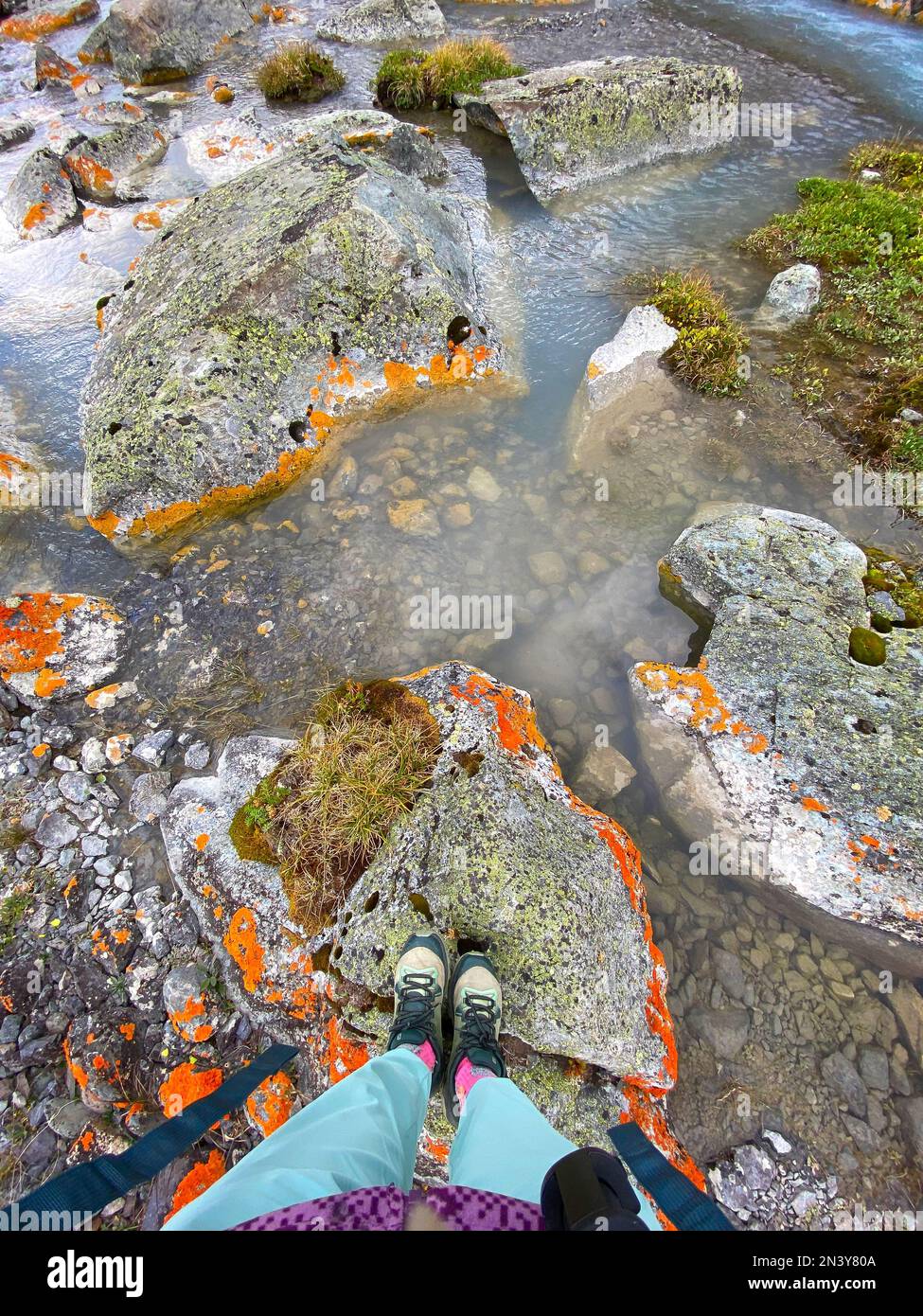 Downward view of the legs and trekking shoes of a traveler girl standing on alpine stones with ...