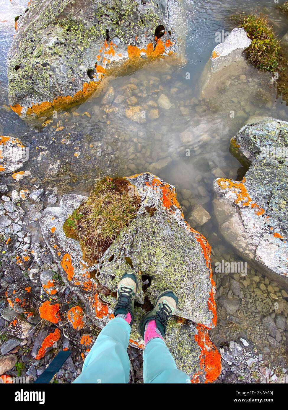 Downward view of the legs and trekking shoes of a traveler girl standing on alpine stones with ...
