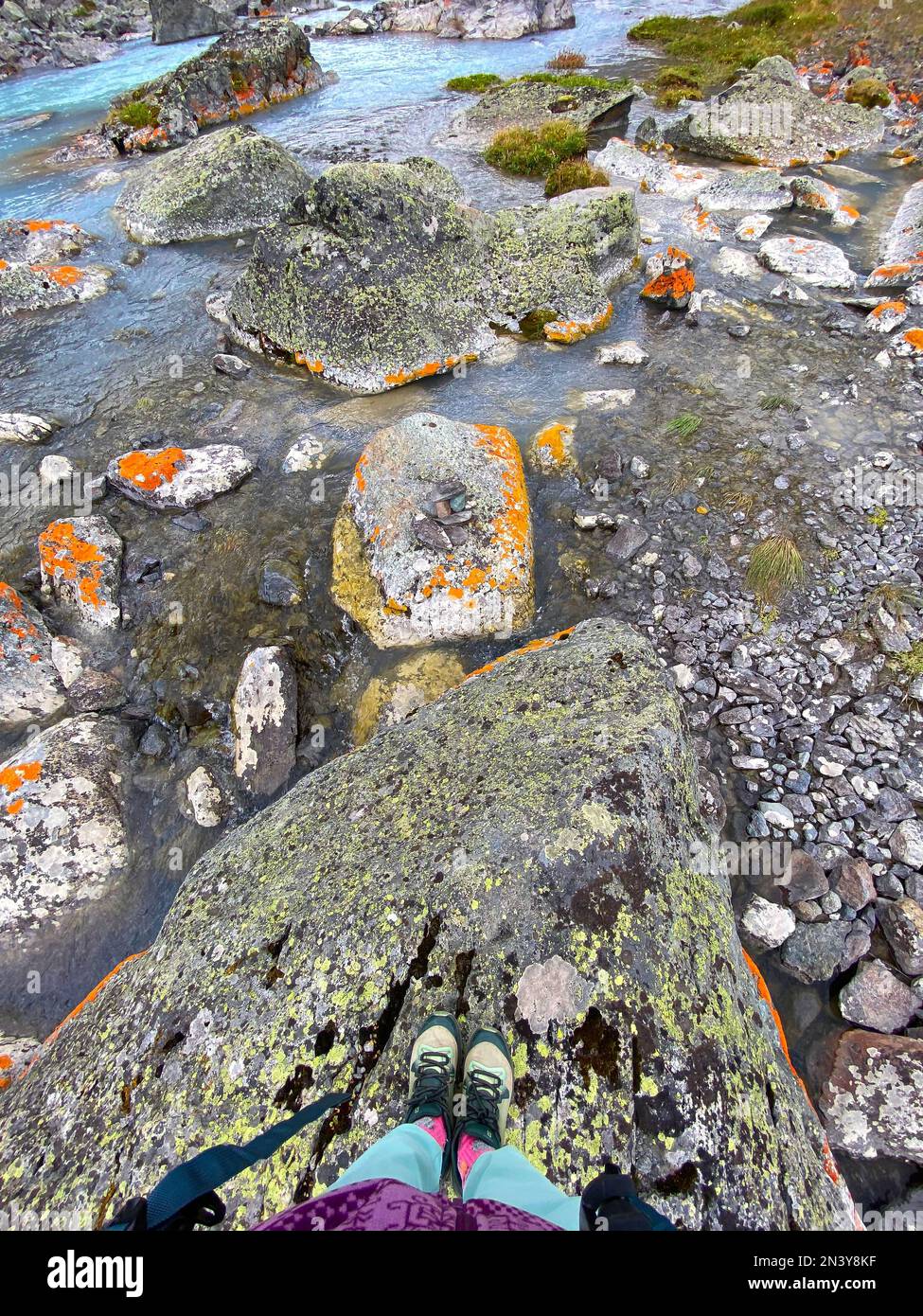 Downward view of the legs and trekking shoes of a traveler girl standing on alpine stones with ...