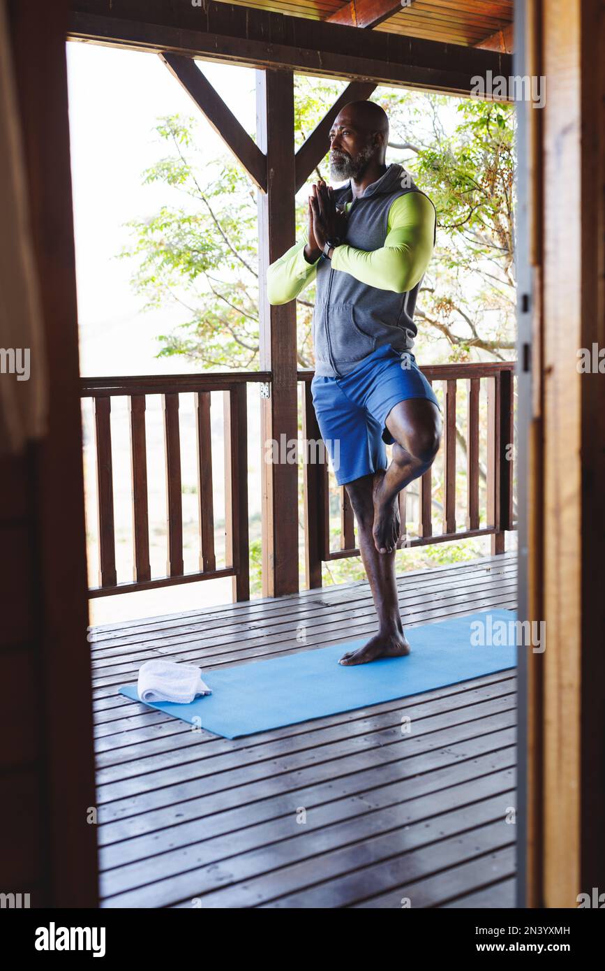 African american bald senior man practicing tree pose in balcony at log cabin seen through ...