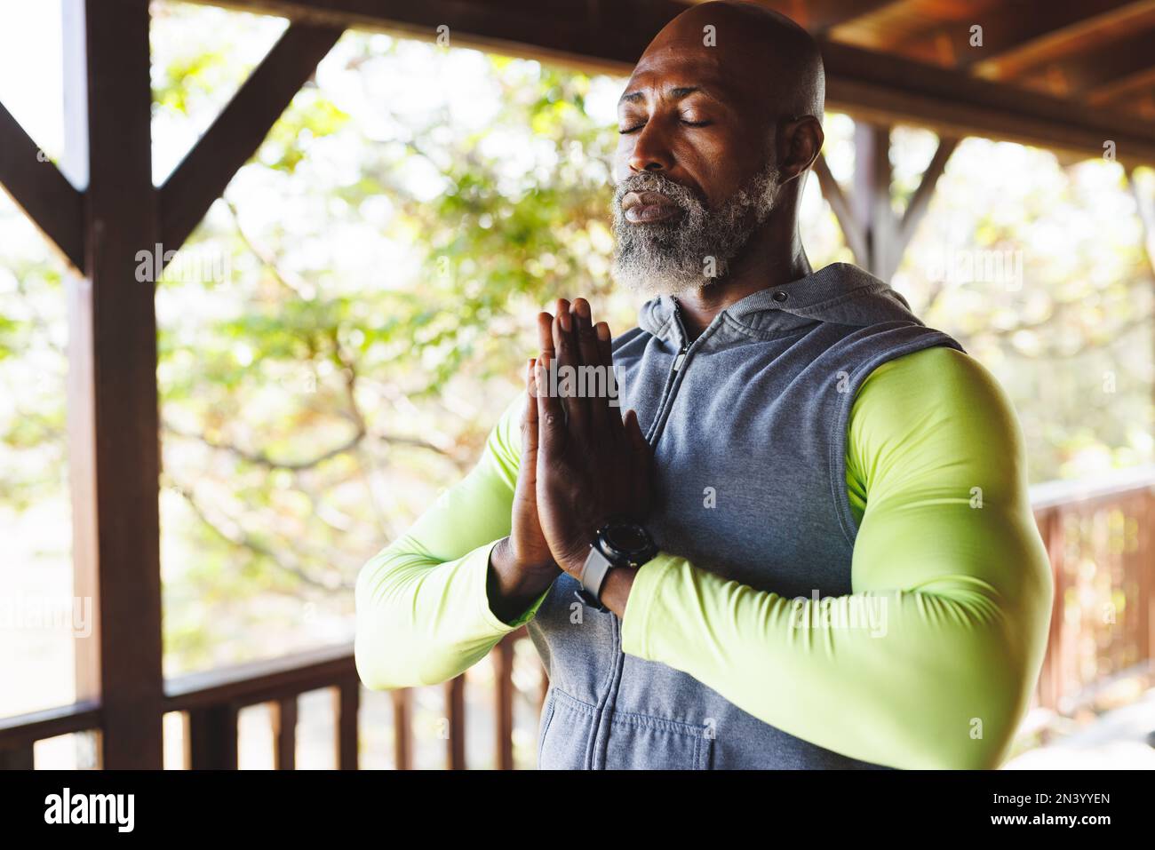 Bald african american senior man meditating in prayer pose while standing in balcony at log ...