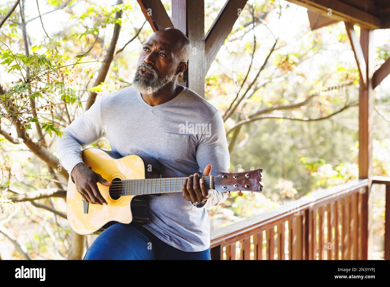 Bald african american senior man playing guitar while sitting on railing in balcony at log cabin ...