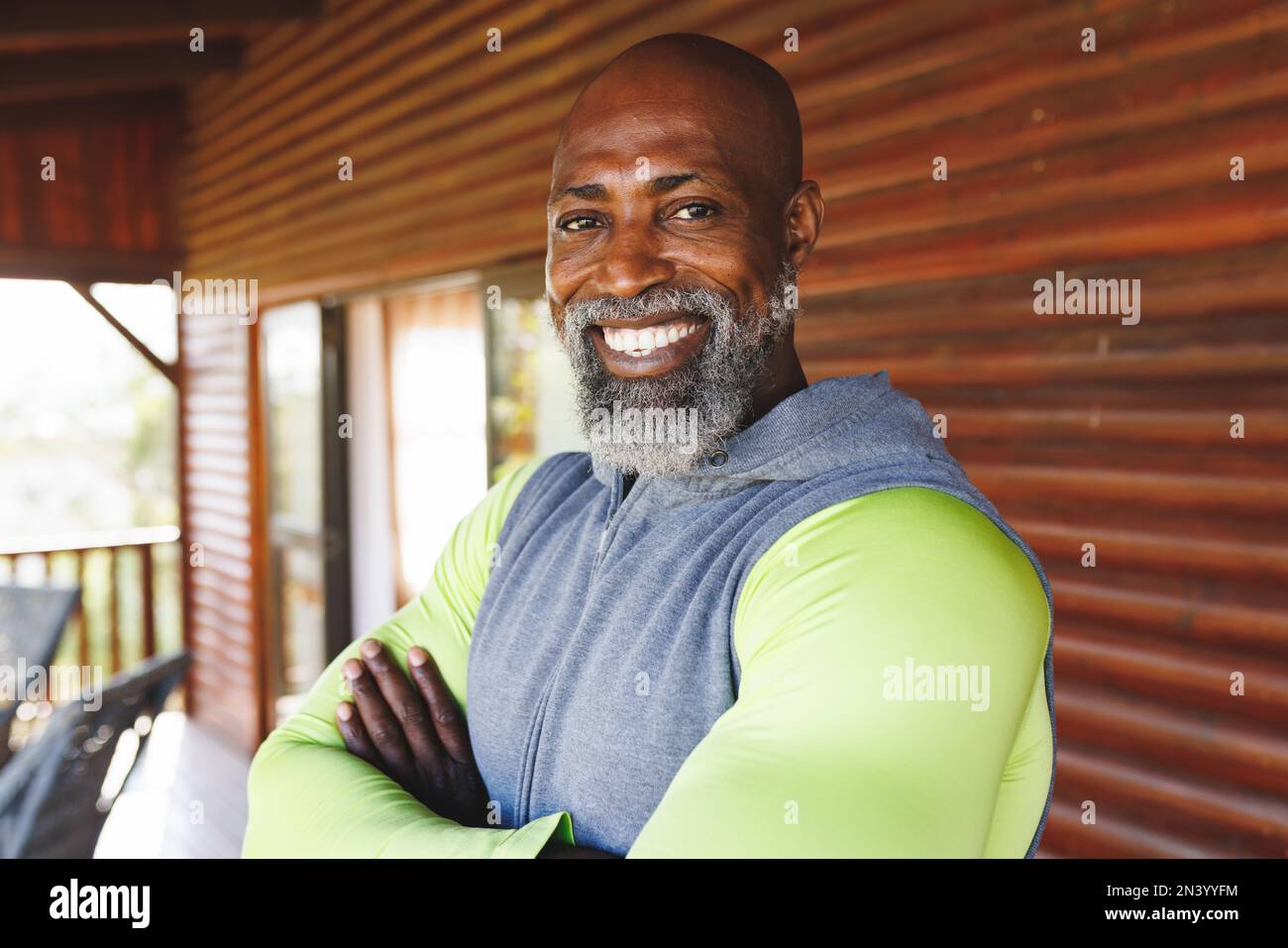 Smiling bald african american senior man with arms crossed standing against wall in log cabin ...