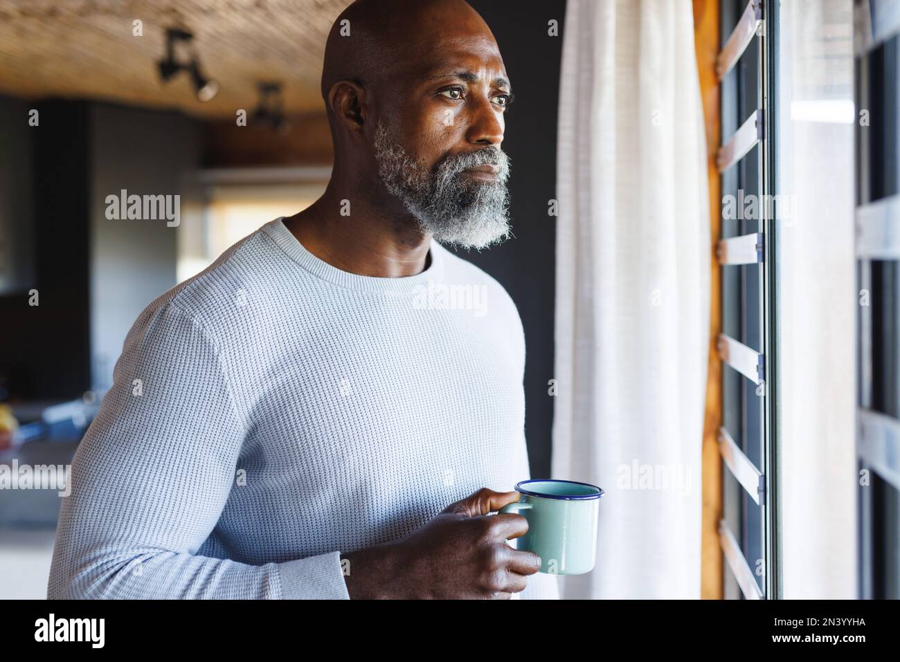 Thoughtful bald african american senior man with coffee mug looking through window at log cabin ...
