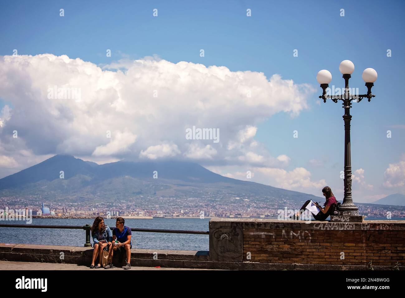 Naples, Italy - Young girl sat on a wall, leaning over a lamp post and ...