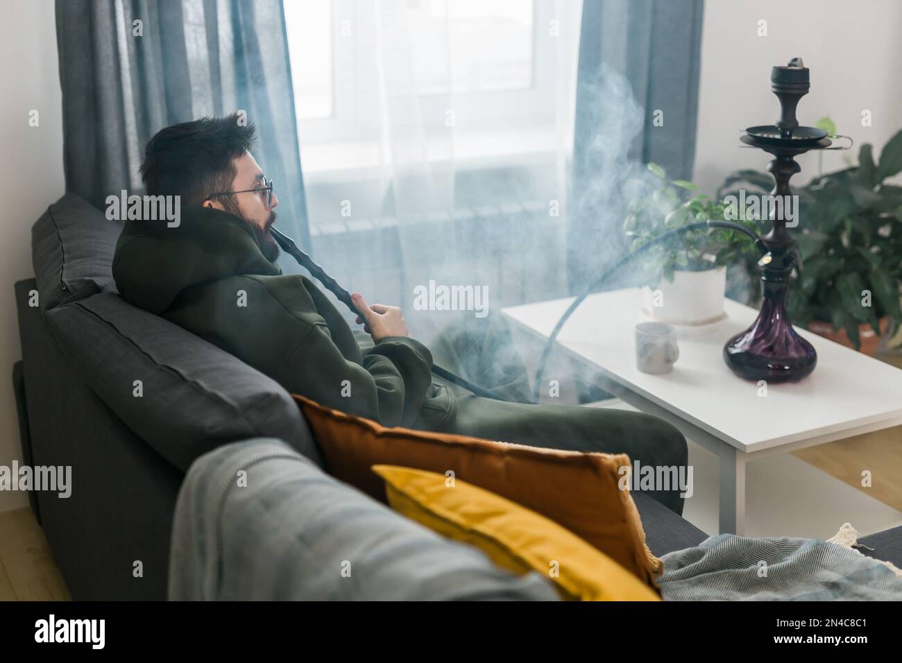 Bearded millennial or gen z man smoking hookah while relaxing on sofa ...