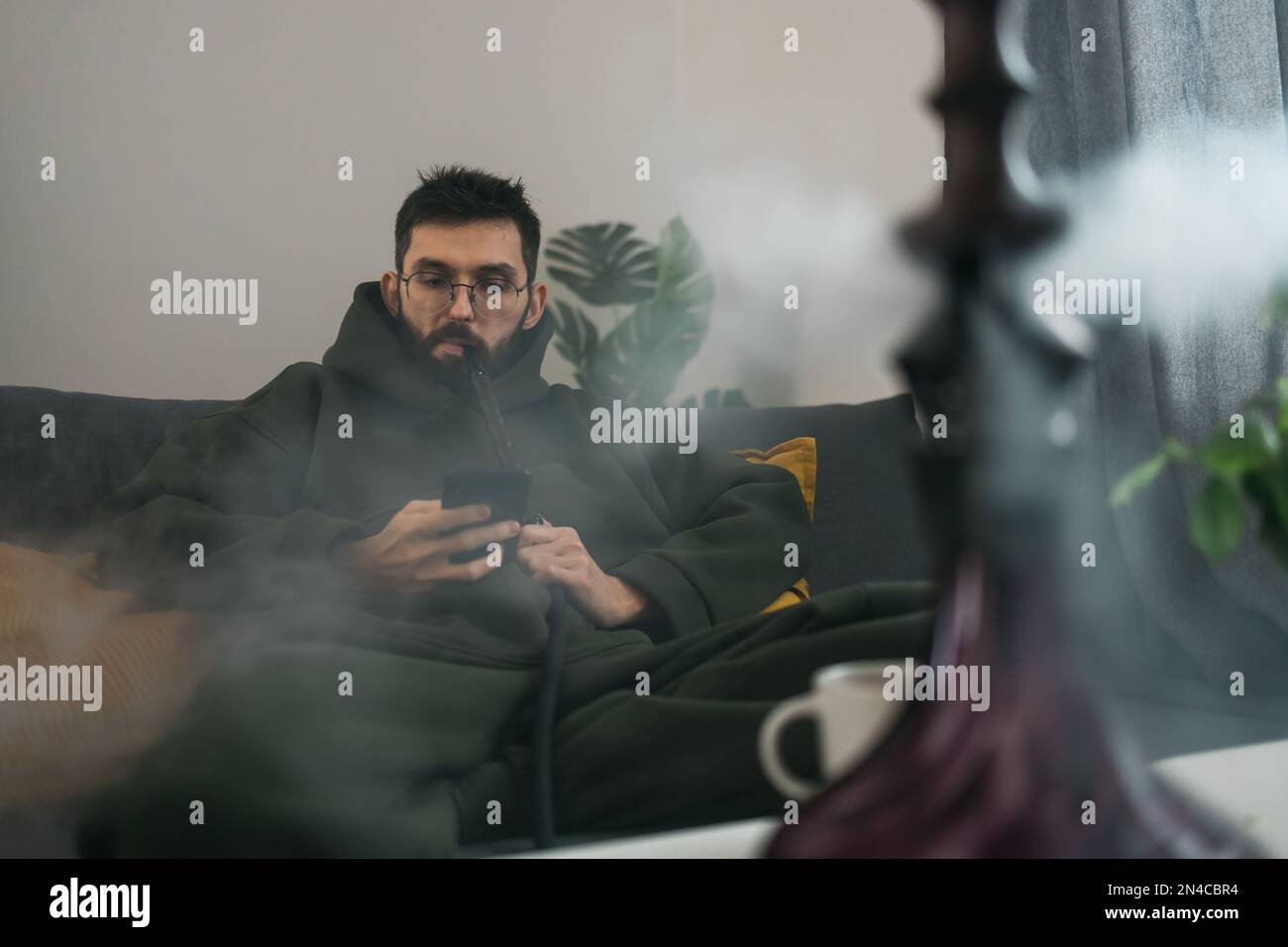 Bearded millennial or gen z man smoking hookah while relaxing on sofa ...
