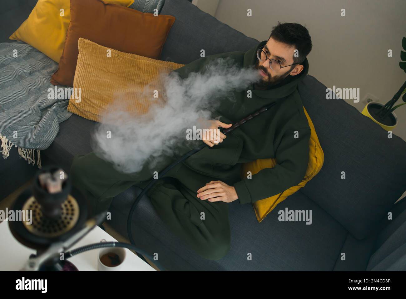 Bearded millennial or gen z man smoking hookah while relaxing on sofa ...