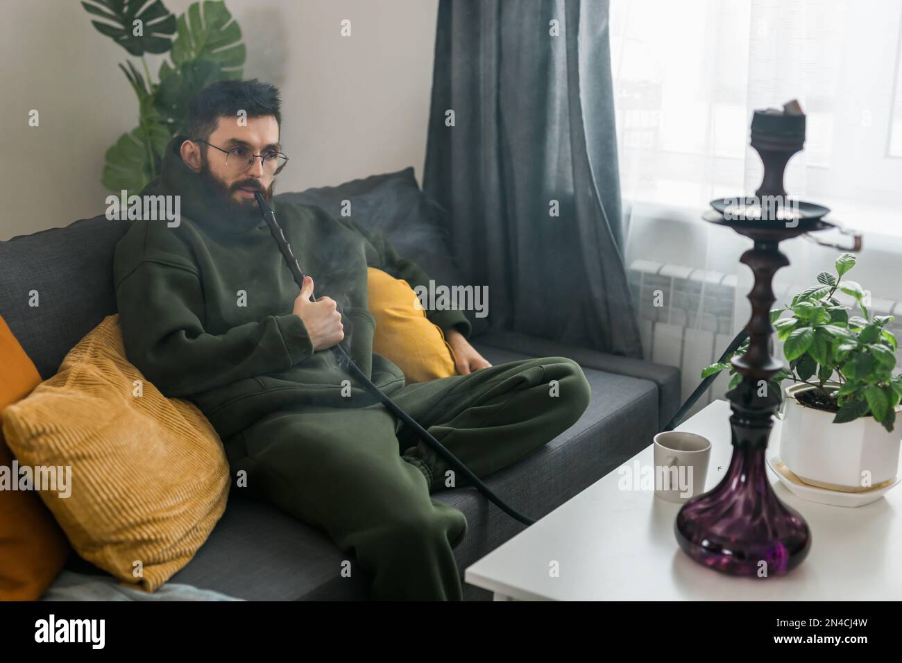 Bearded millennial or gen z man smoking hookah while relaxing on sofa ...