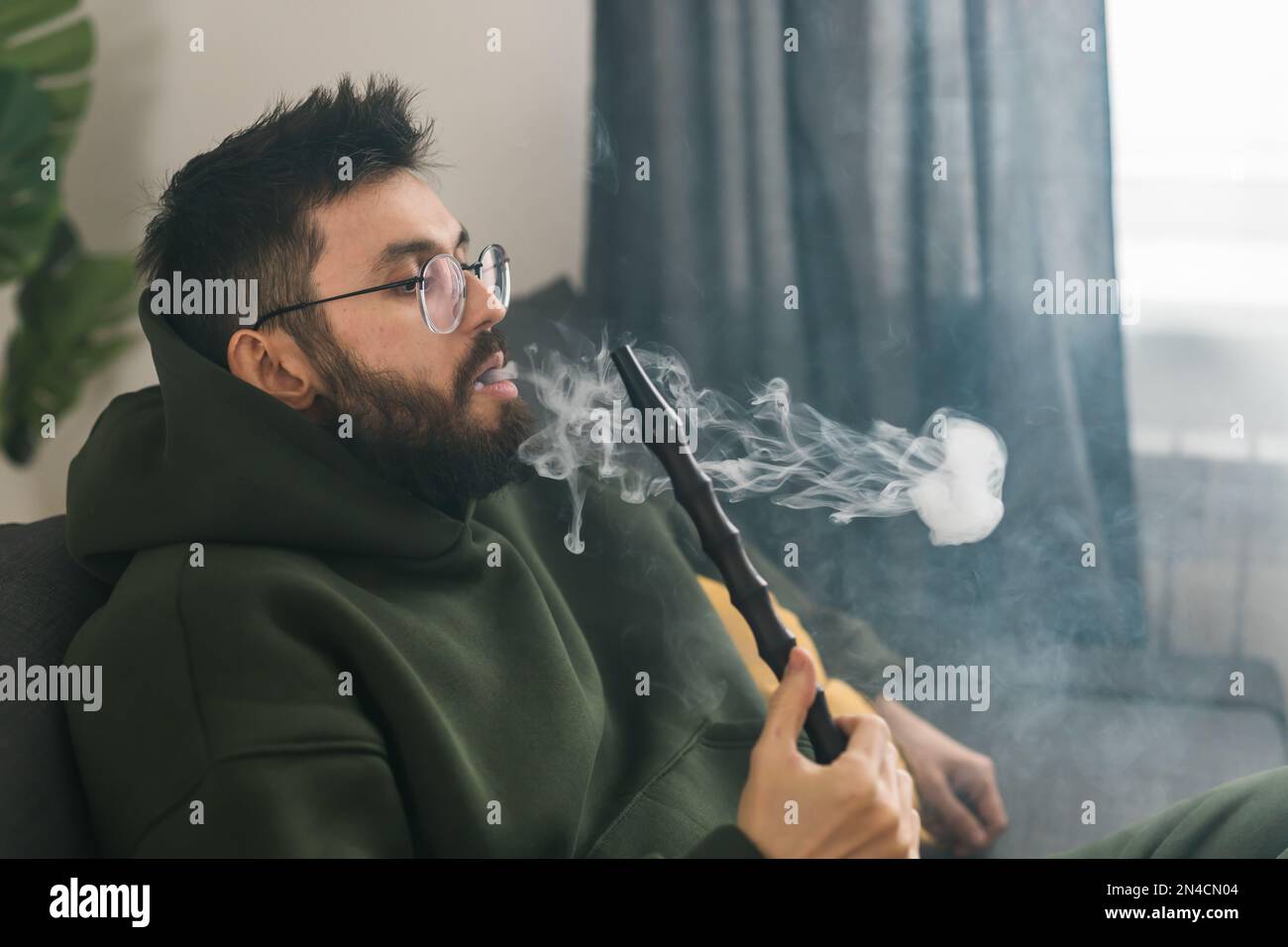 Bearded millennial or gen z man smoking hookah while relaxing on sofa ...