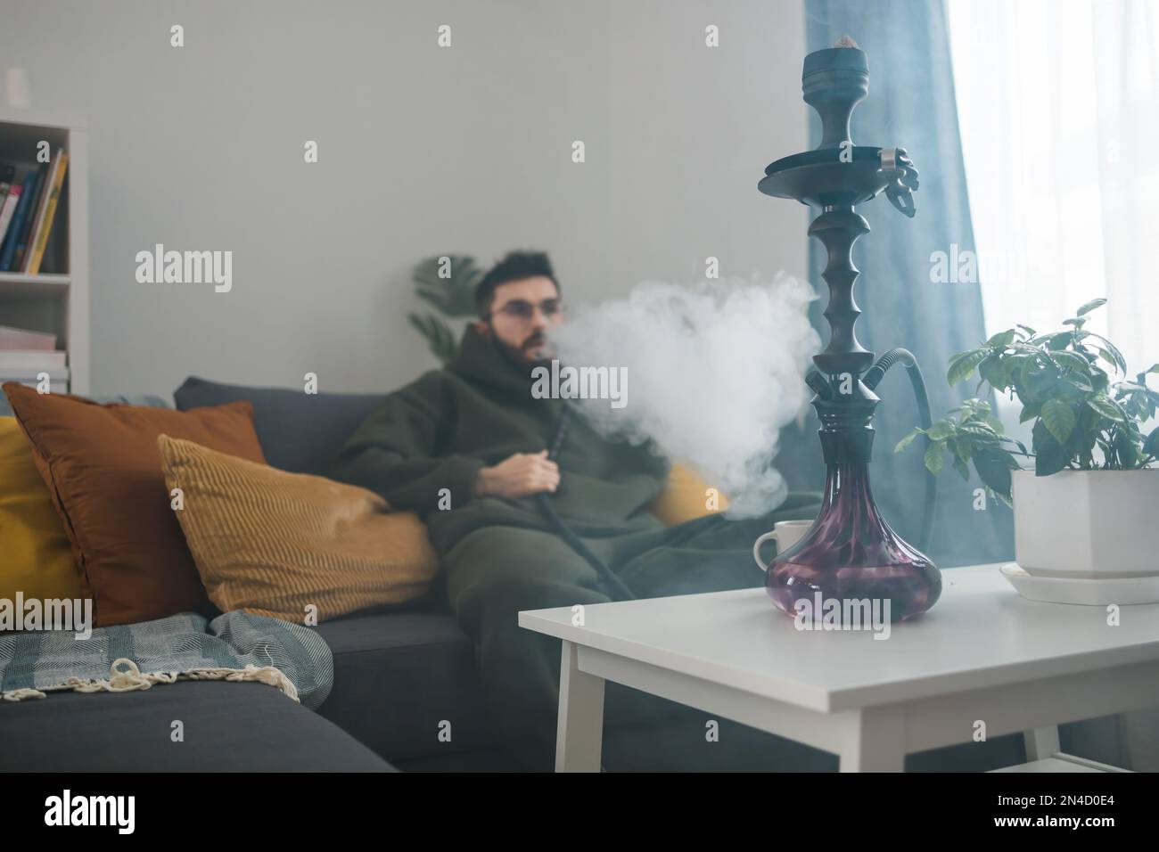 Bearded millennial or gen z man smoking hookah while relaxing on sofa ...
