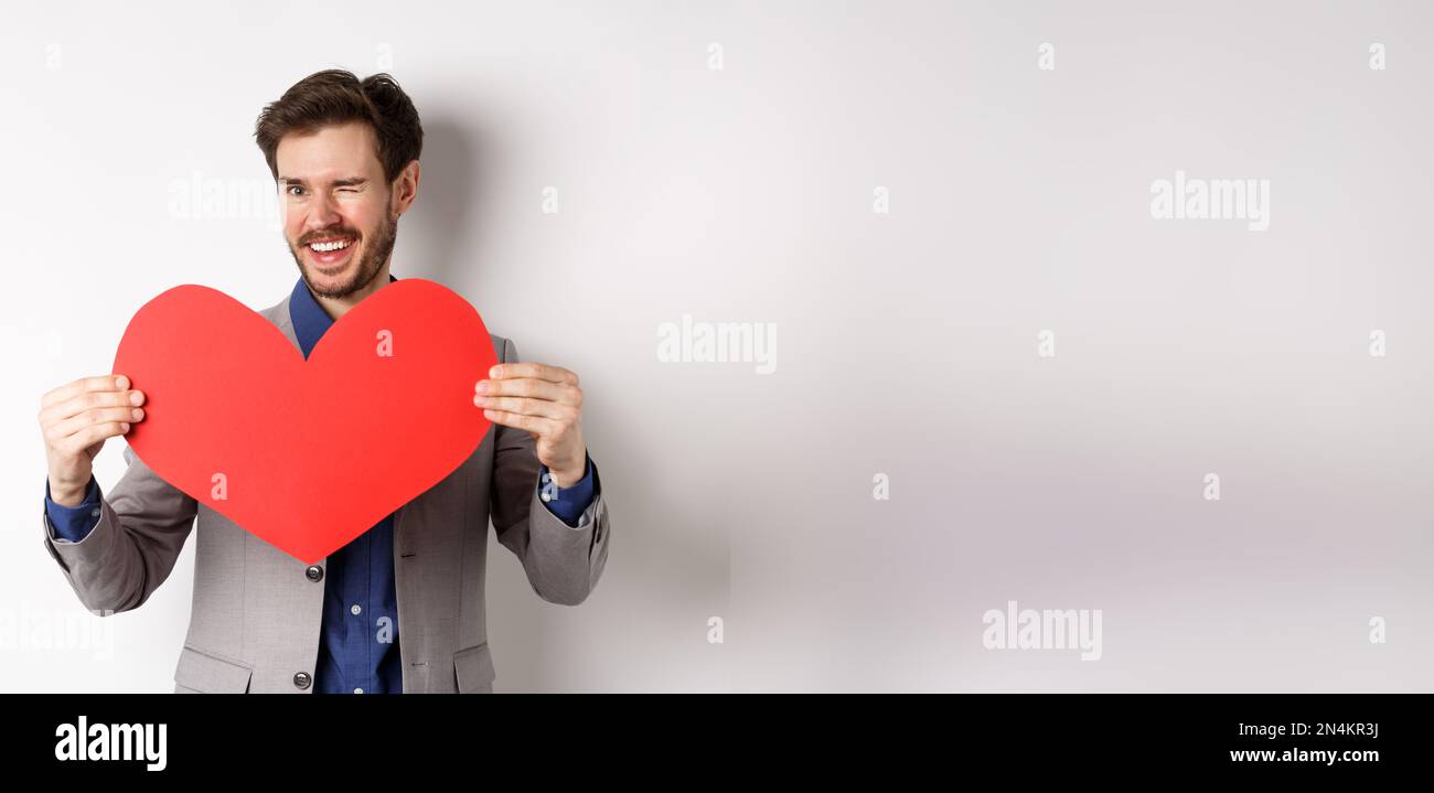 Charismatic young man winking and smiling, showing big red heart cutout ...