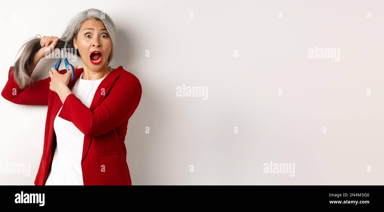 Scared asian senior woman cutting hair with scissors and screaming in ...