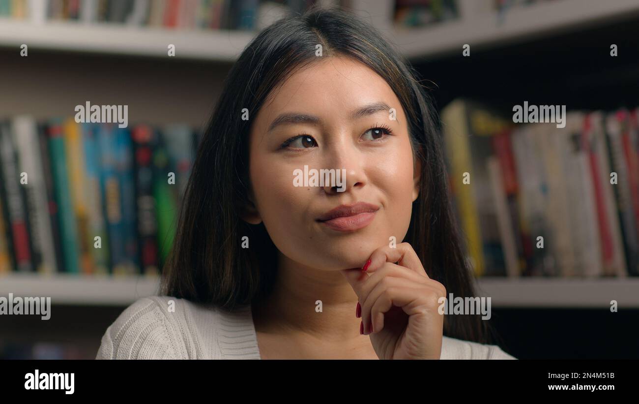 Portrait pensive korean woman standing in library near bookshelf think ...