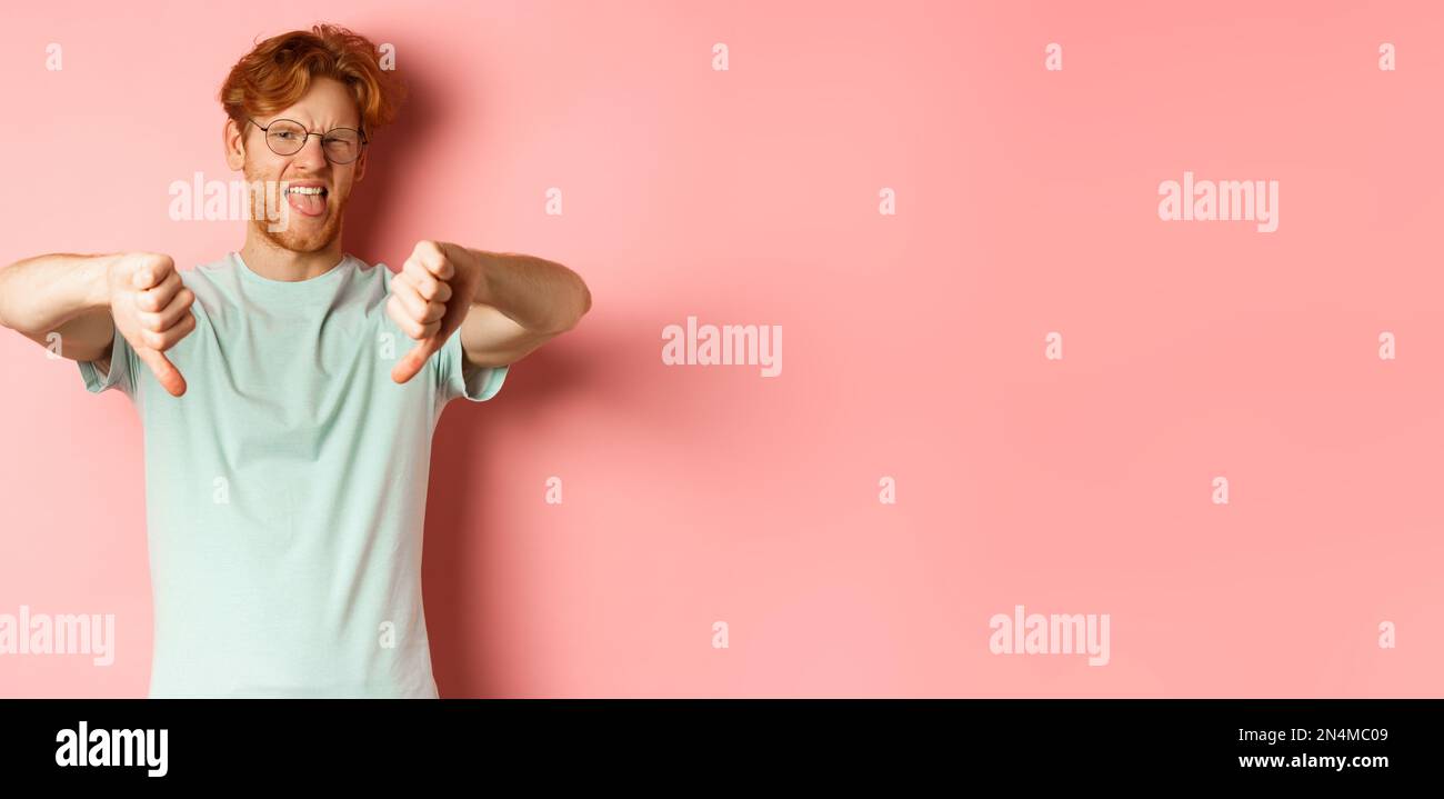 Disgusted redhead man in t-shirt and glasses showing thumbs down and ...