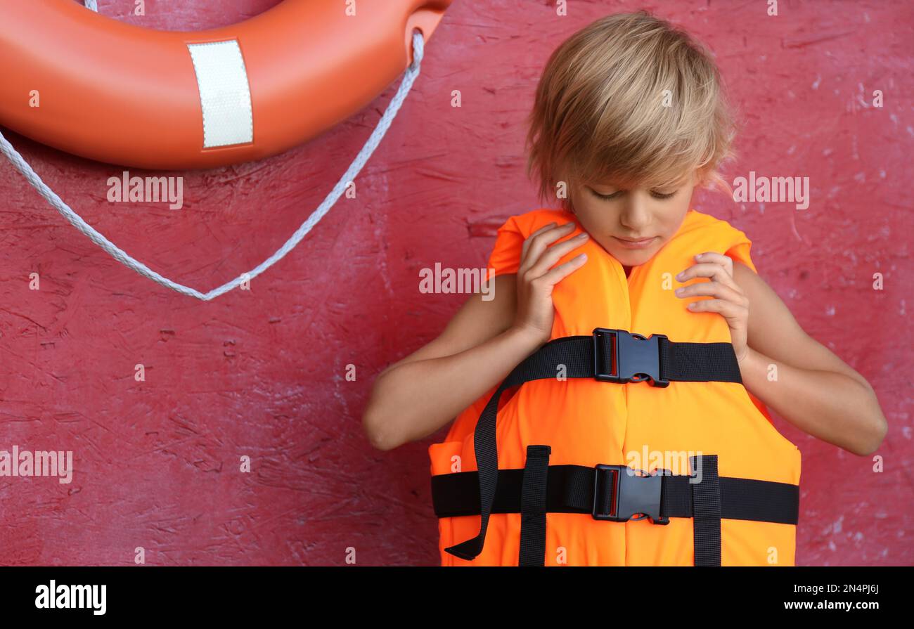 Little boy putting on orange life vest near red wall with safety ring ...