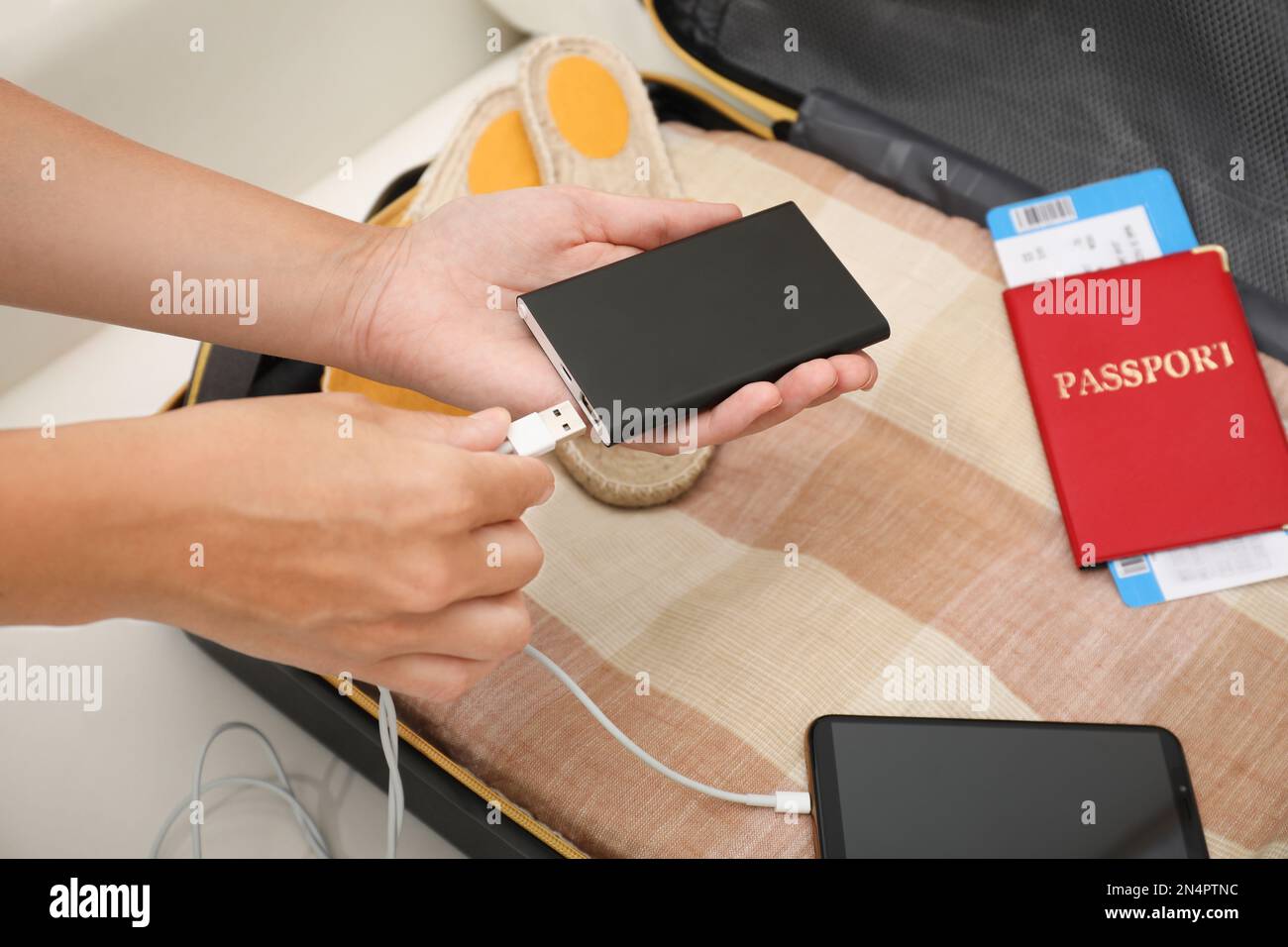 Woman charging smartphone with power bank near packed suitcase indoors ...